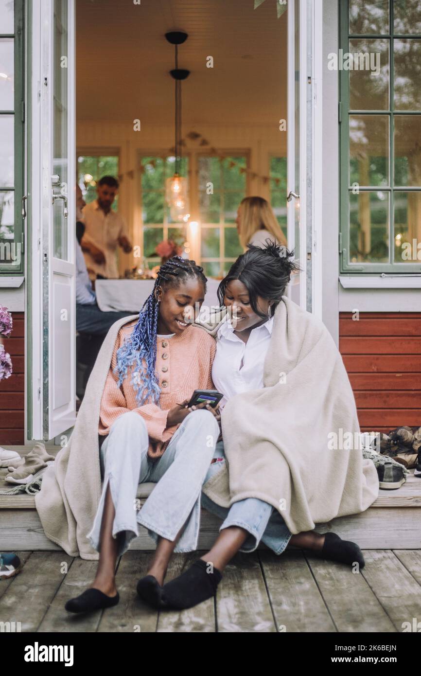 Mother and daughter covered in blanket sharing smart phone while ...