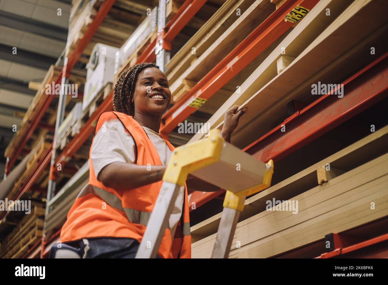 Smiling carpenter standing on ladder by rack while working in warehouse ...