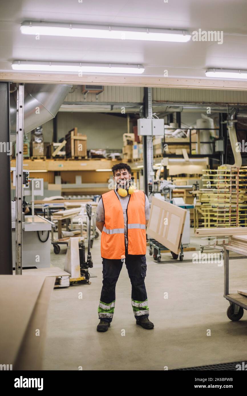 Full length of smiling carpenter wearing reflective clothing and ear ...