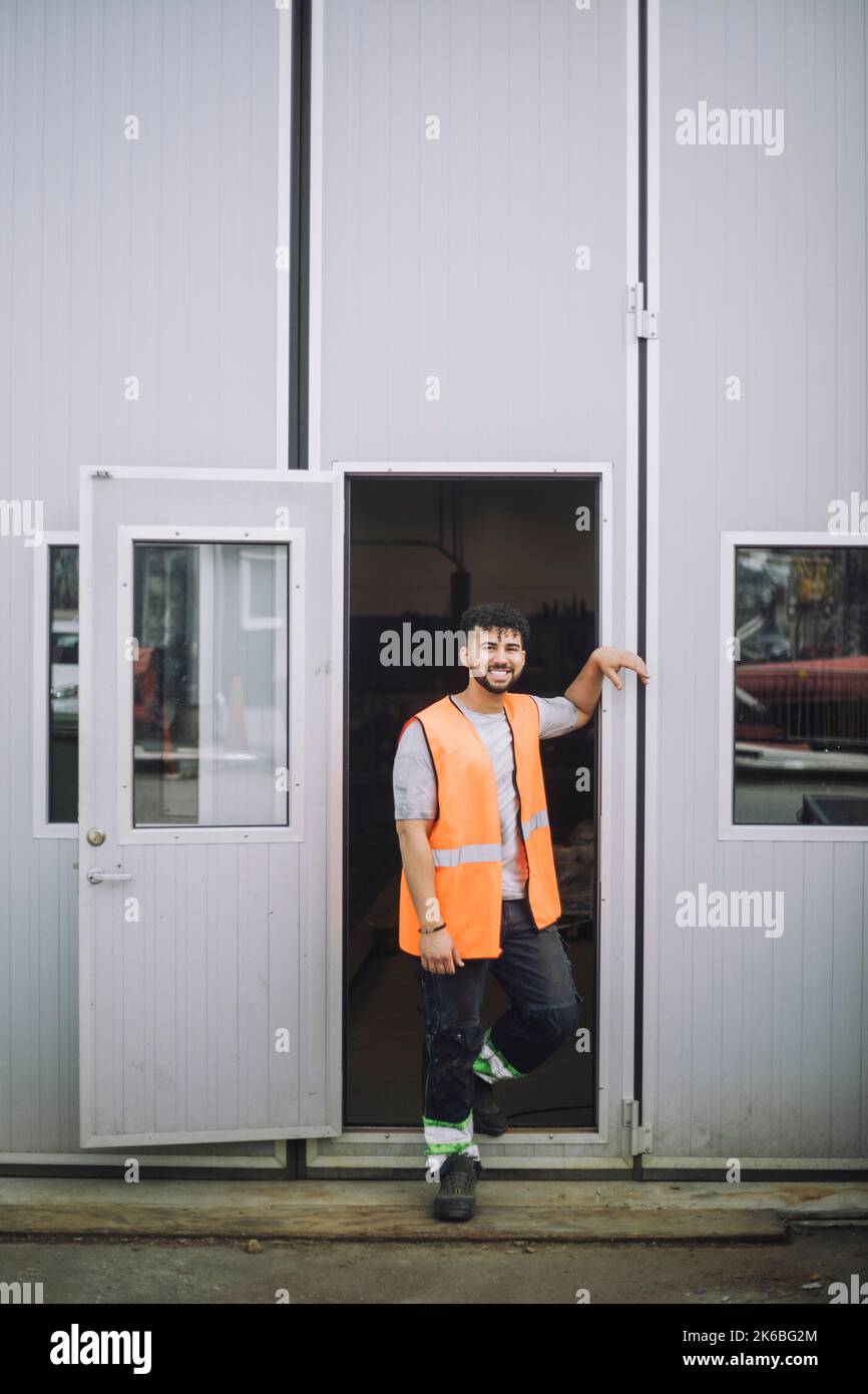 Full length of smiling young carpenter standing at doorway of warehouse ...