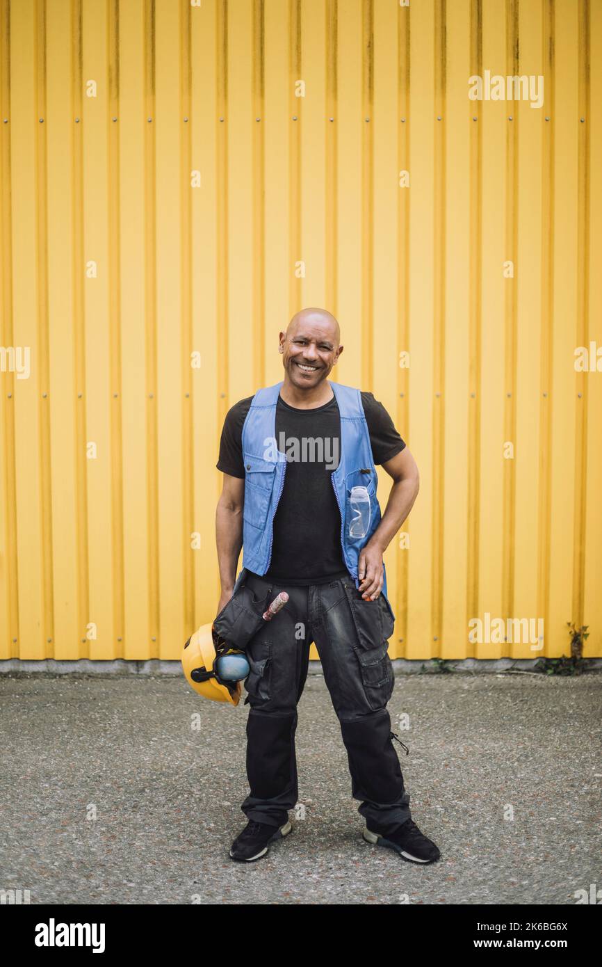 Full length of smiling bald construction worker standing against yellow ...