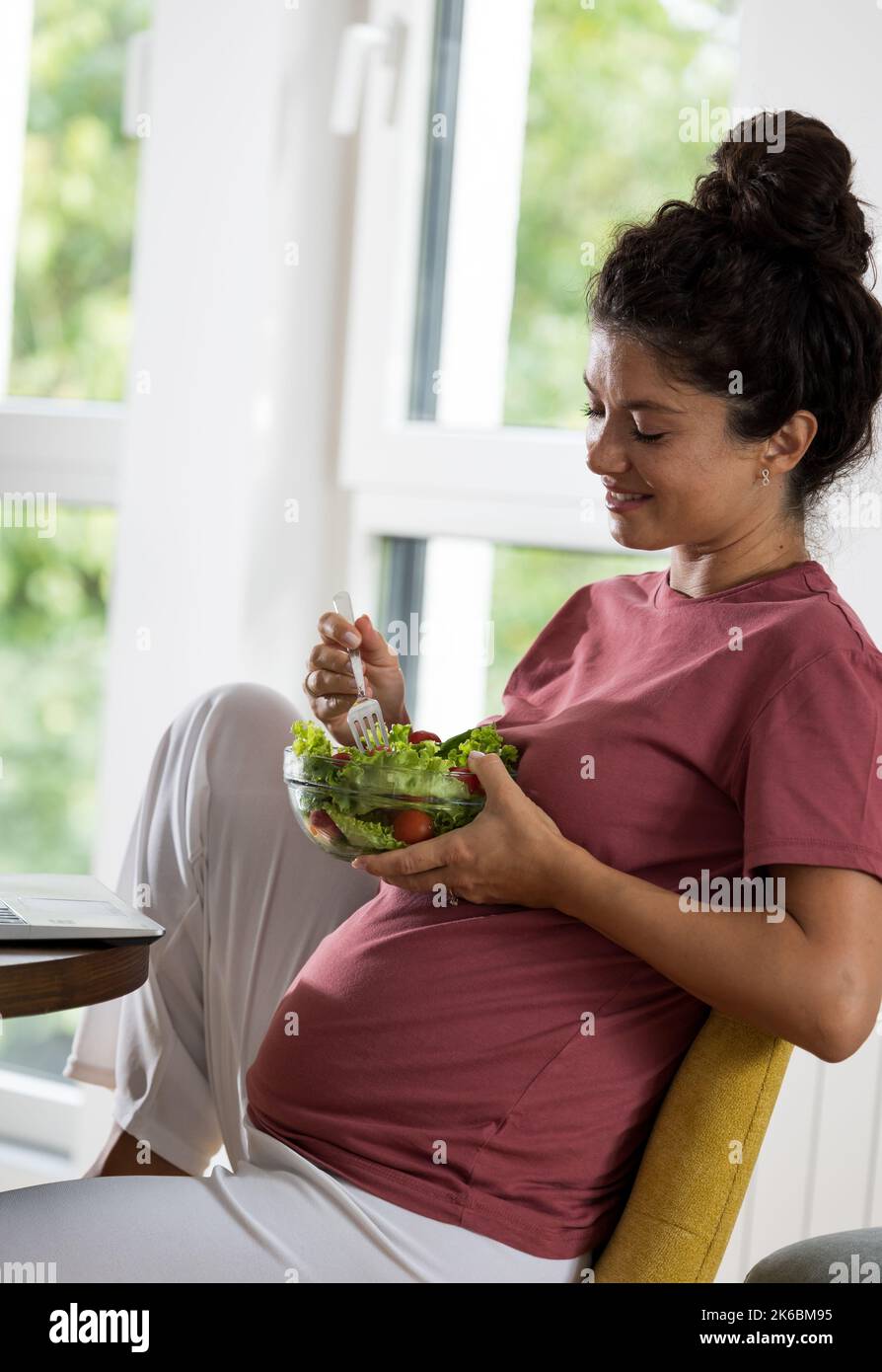 Hungry young woman sitting on dining chair in kitchen and eating salad ...