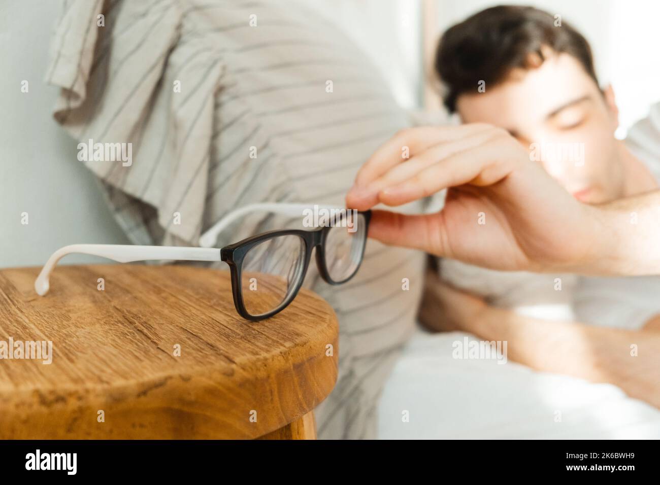A young man reaches for his glasses after waking up from a dream in bed ...
