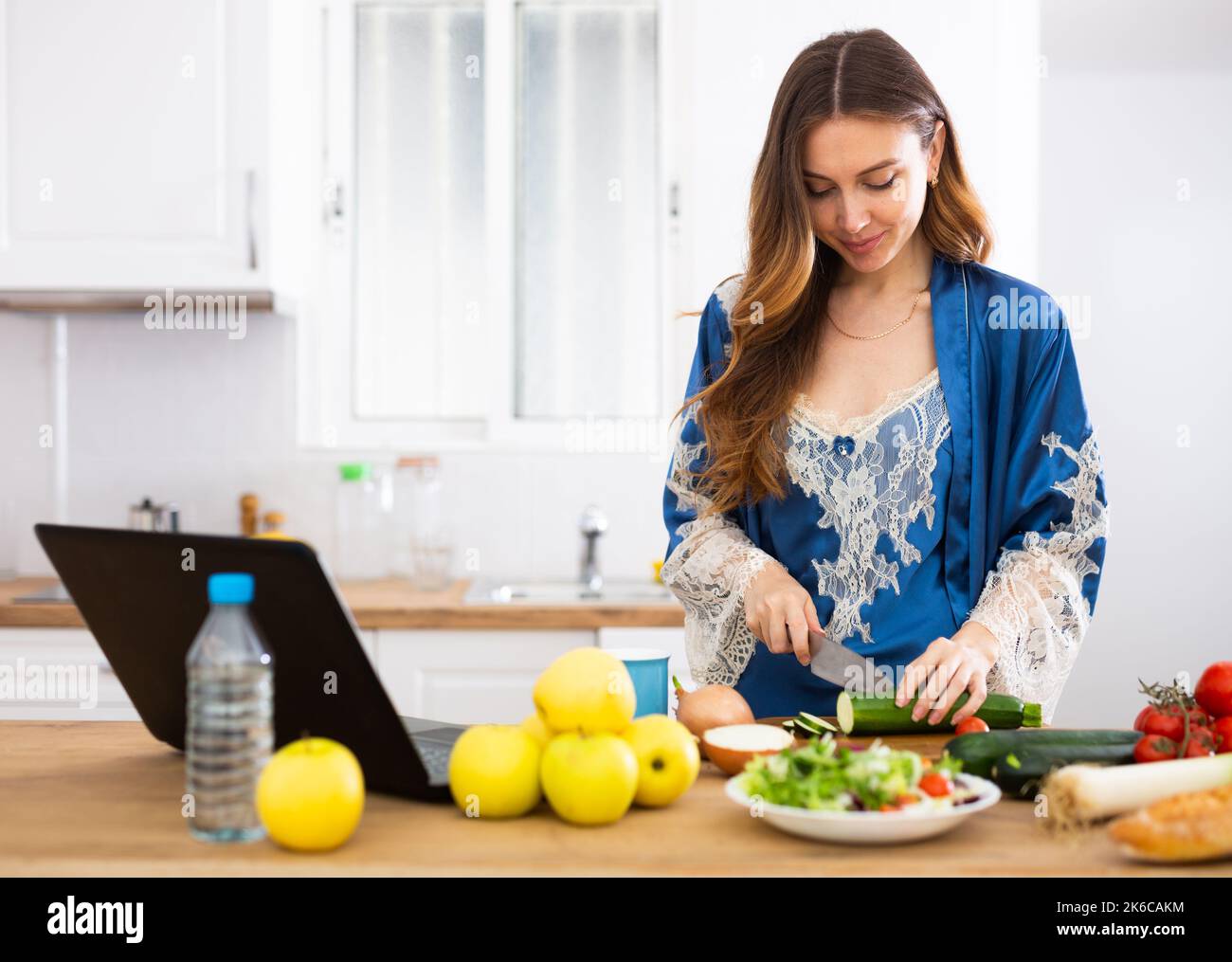 Young woman in blue robe cooking and watching TV series on laptop Stock ...