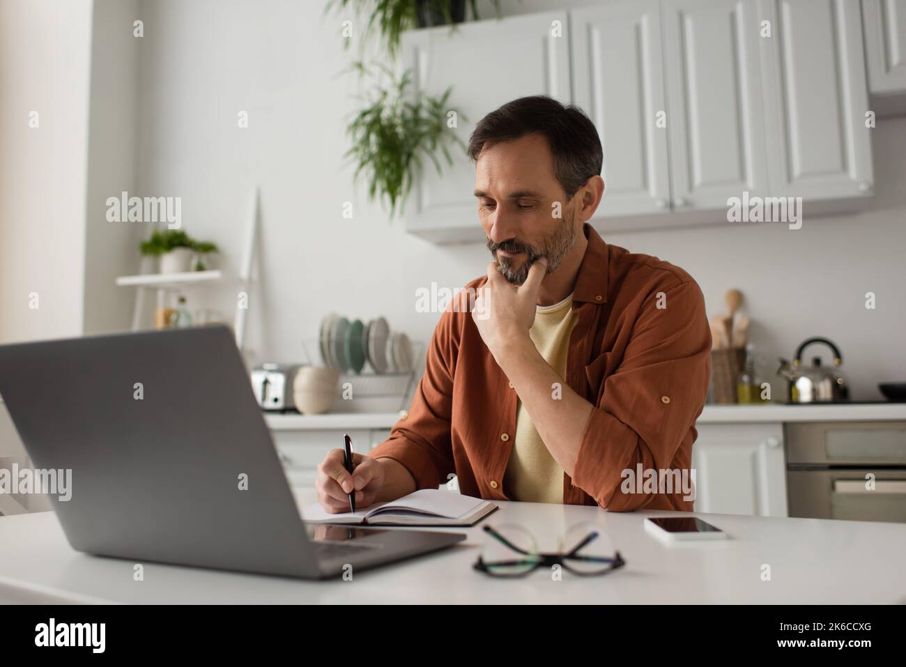 thoughtful man holding hand near face and writing in notebook near ...