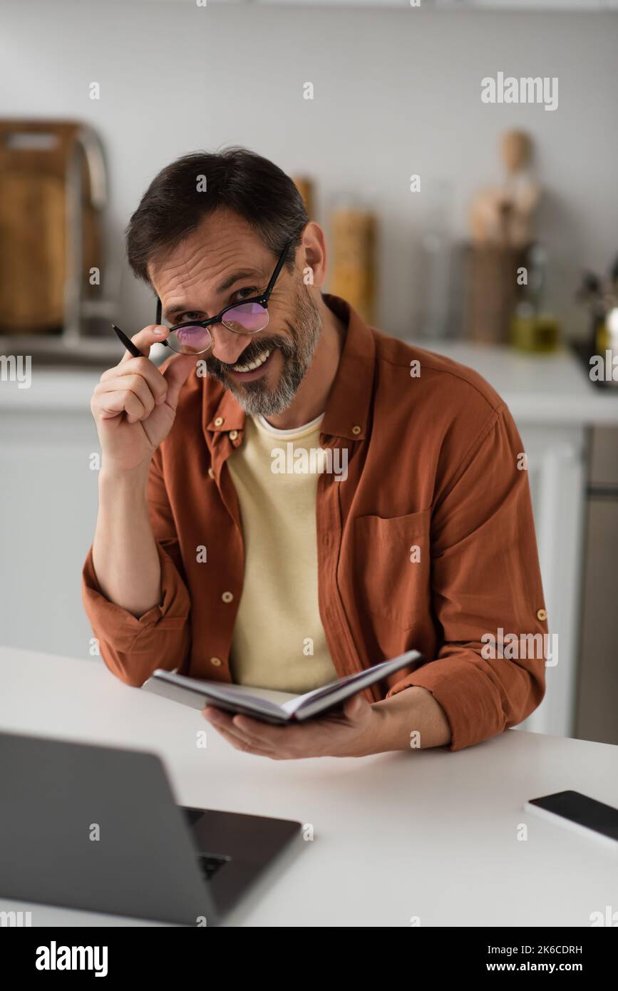 cheerful man in eyeglasses holding notebook and pen while smiling at ...