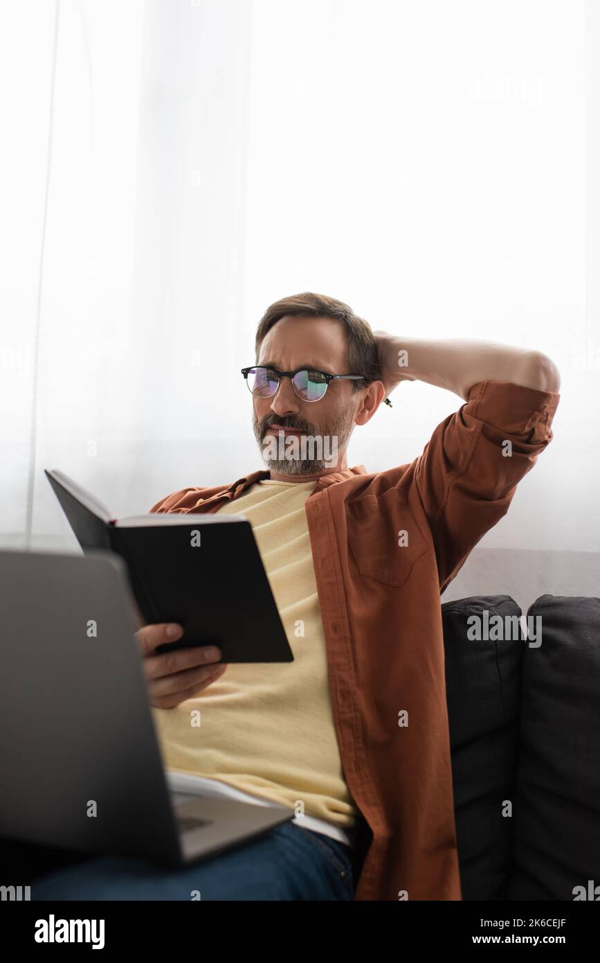pensive man in eyeglasses looking in notebook while sitting on couch ...