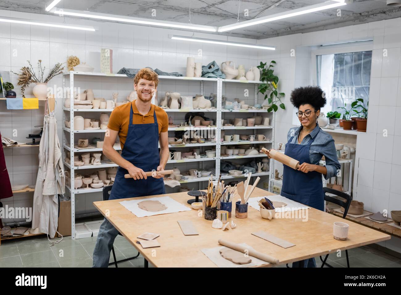 happy interracial couple in aprons holding rolling pins in pottery ...