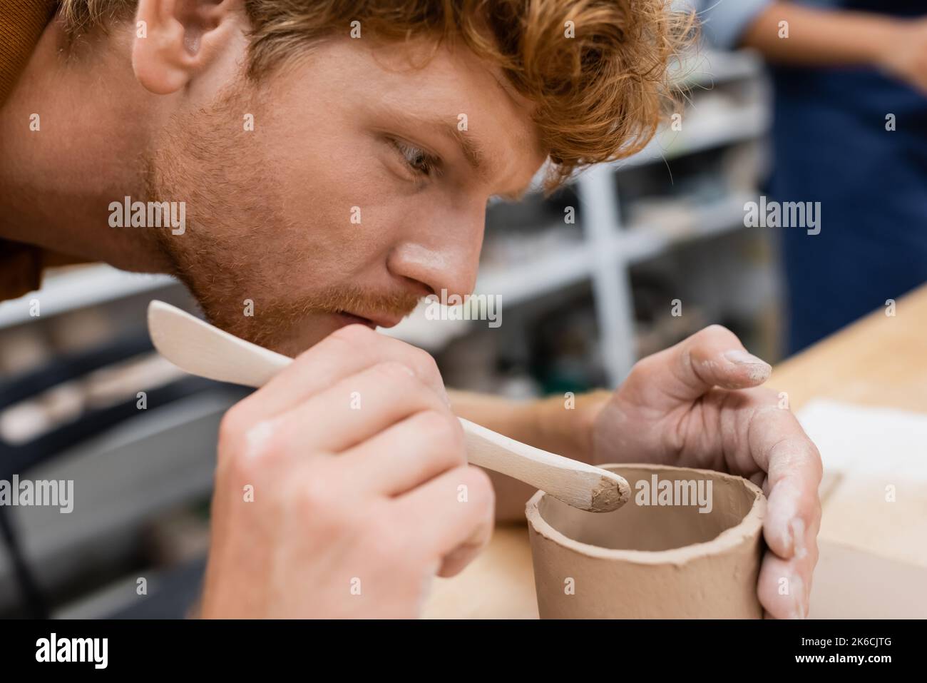 concentrated man with red hair holding shaper while modeling clay cup ...