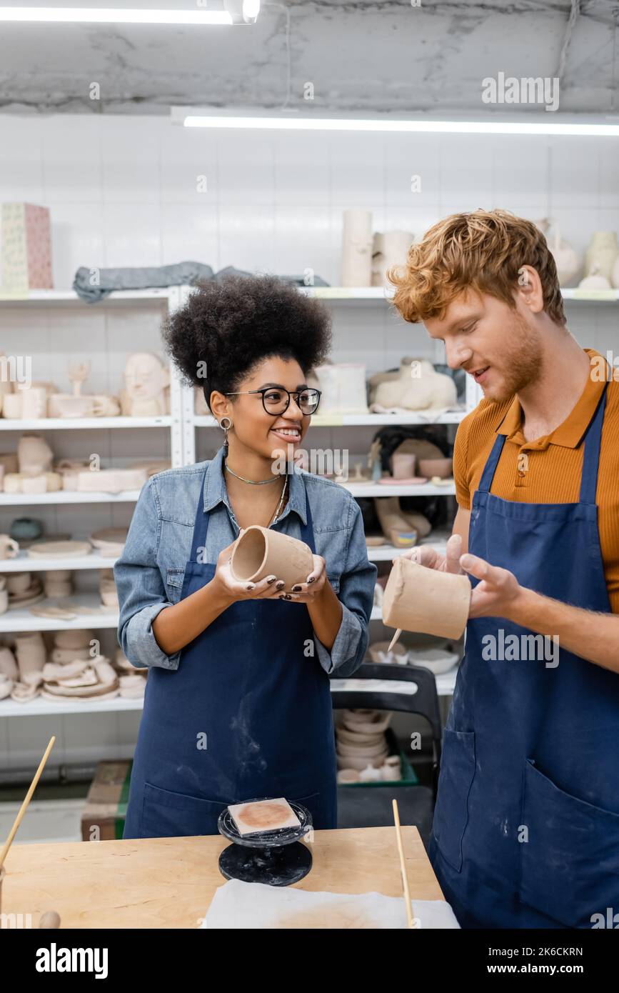 cheerful interracial couple molding clay cups during date in pottery ...