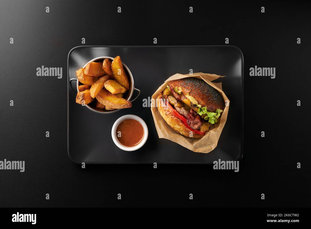 A top view of a burger near fried potato in a bucket on a black plate ...