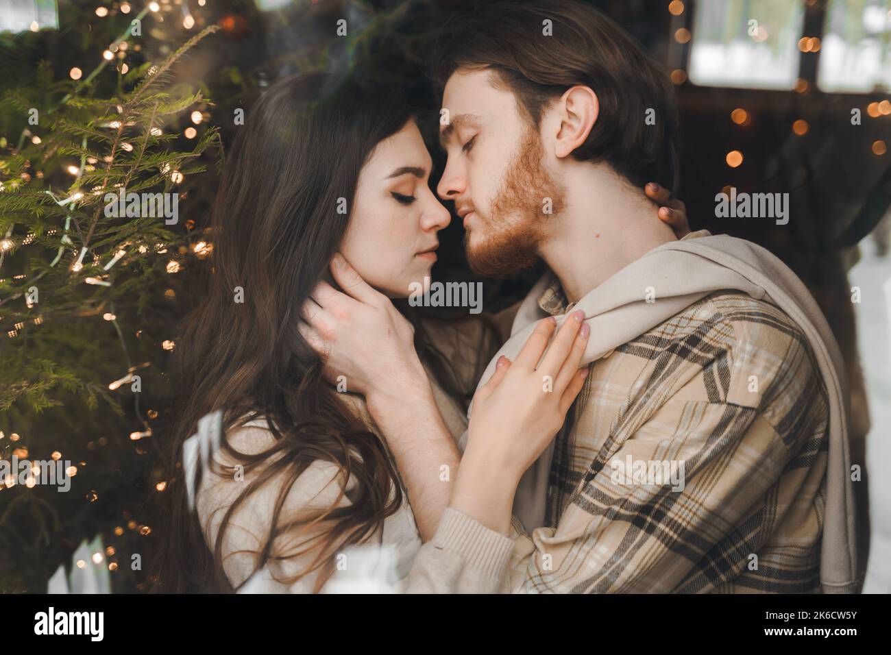 Hugging kissing love couple girl, guy near christmas tree with garland behind window. Lovestory ...
