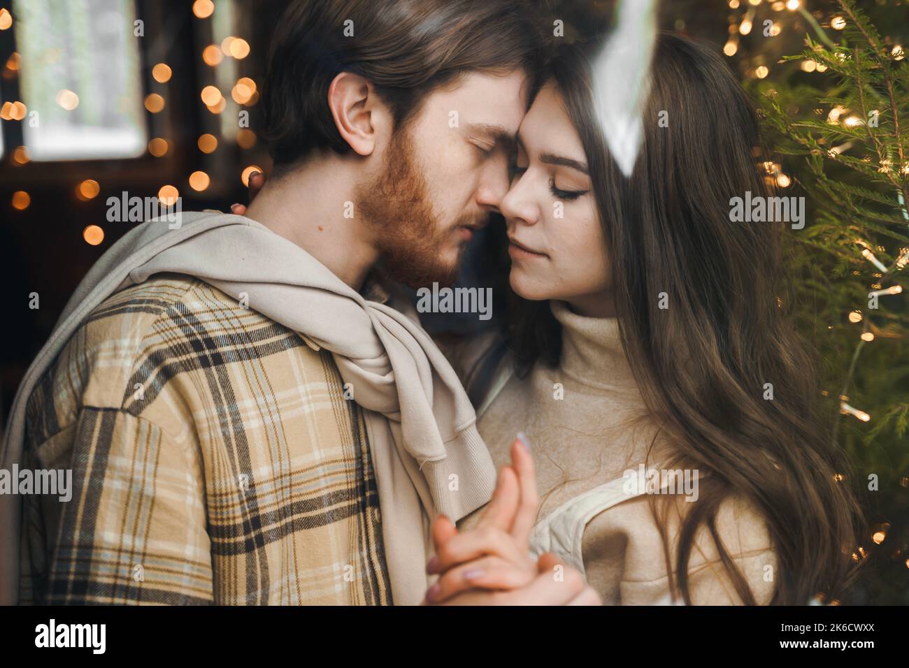 Hugging kissing love couple girl, guy near christmas tree with garland behind window. Lovestory ...