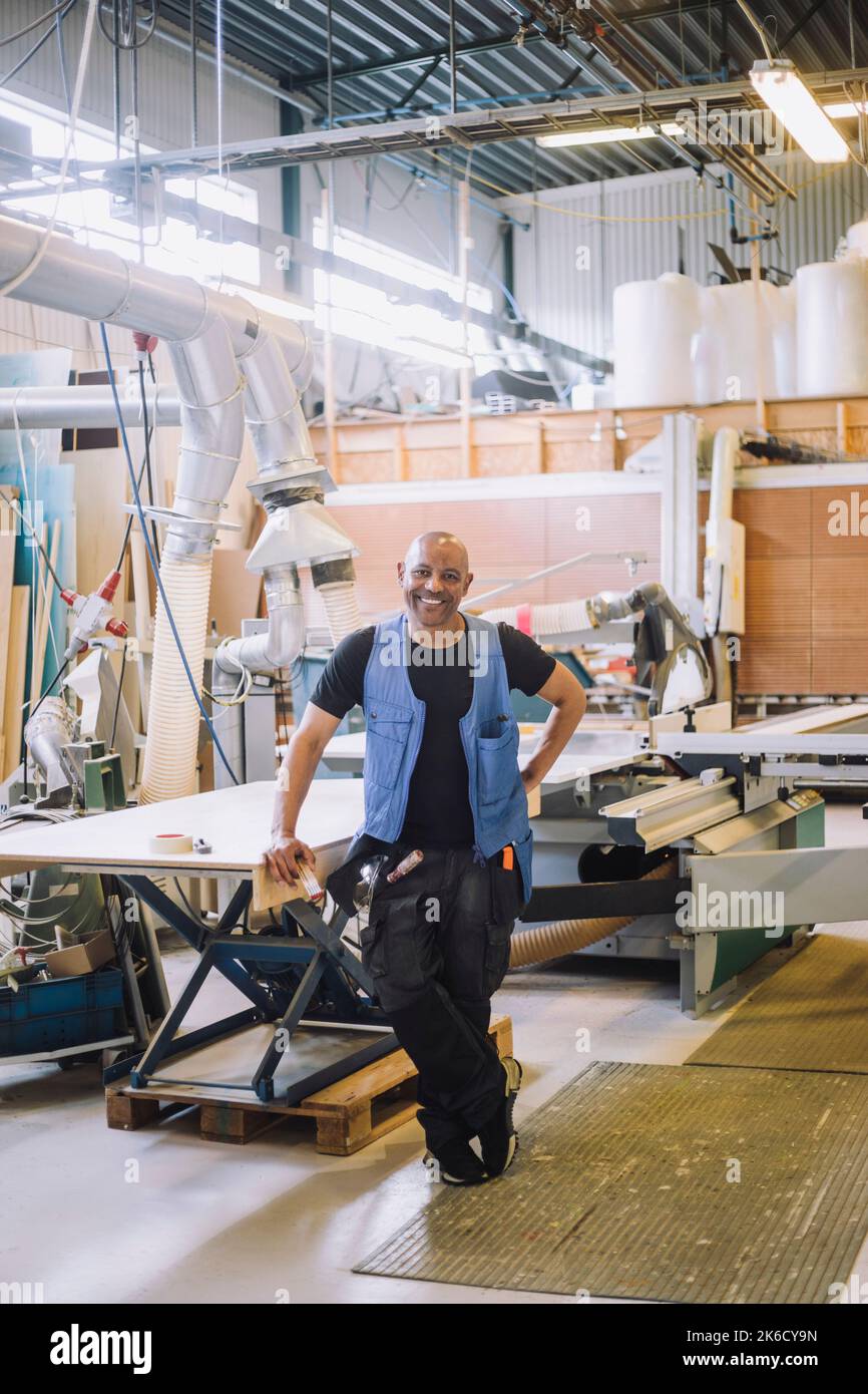Full length of smiling male carpenter standing with hand on hip by ...