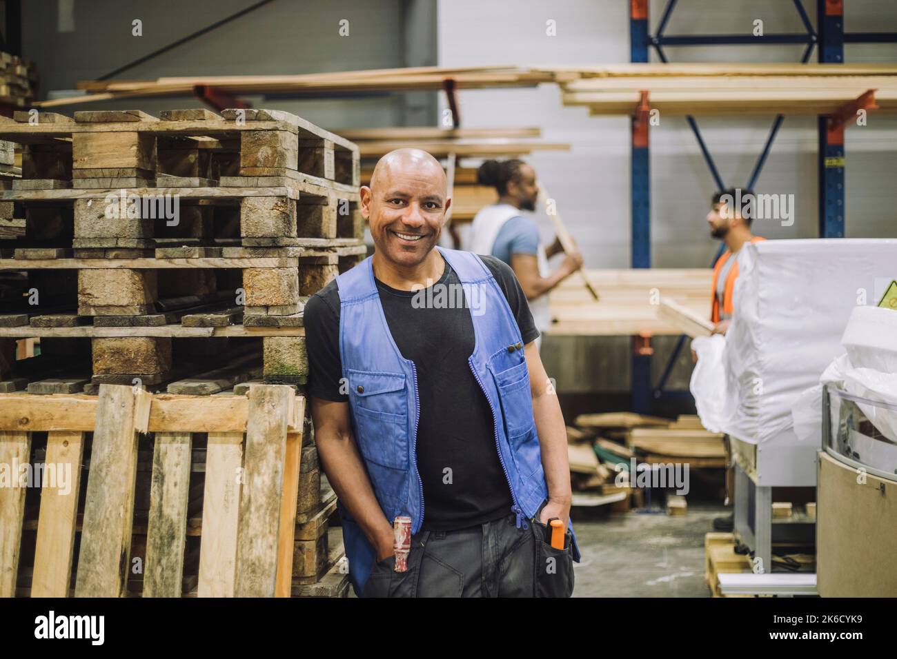 Smiling bald carpenter with hands in pockets leaning on wooden rack in ...