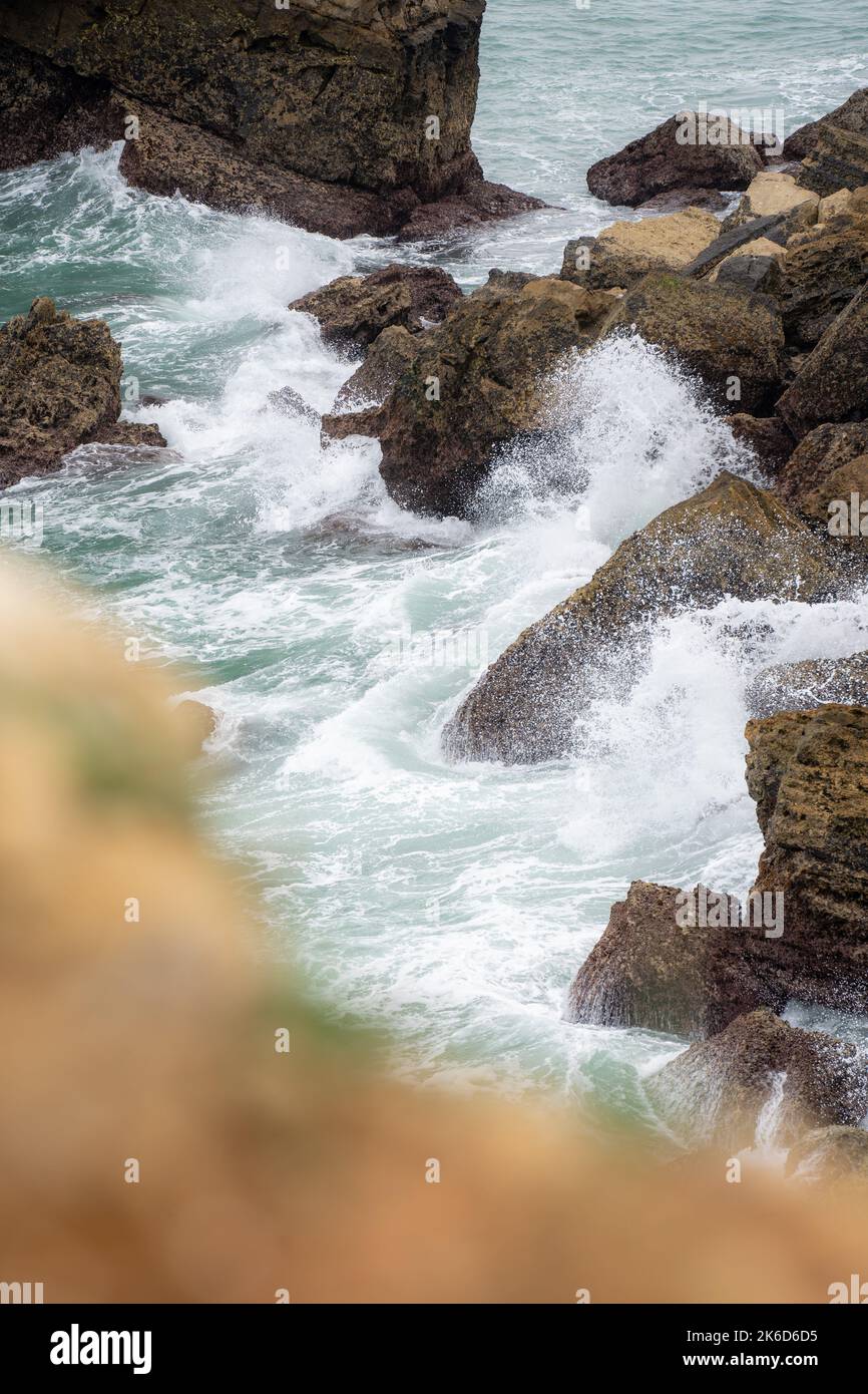 A vertical shot of foamy ocean waves hitting the big coastal rocks ...