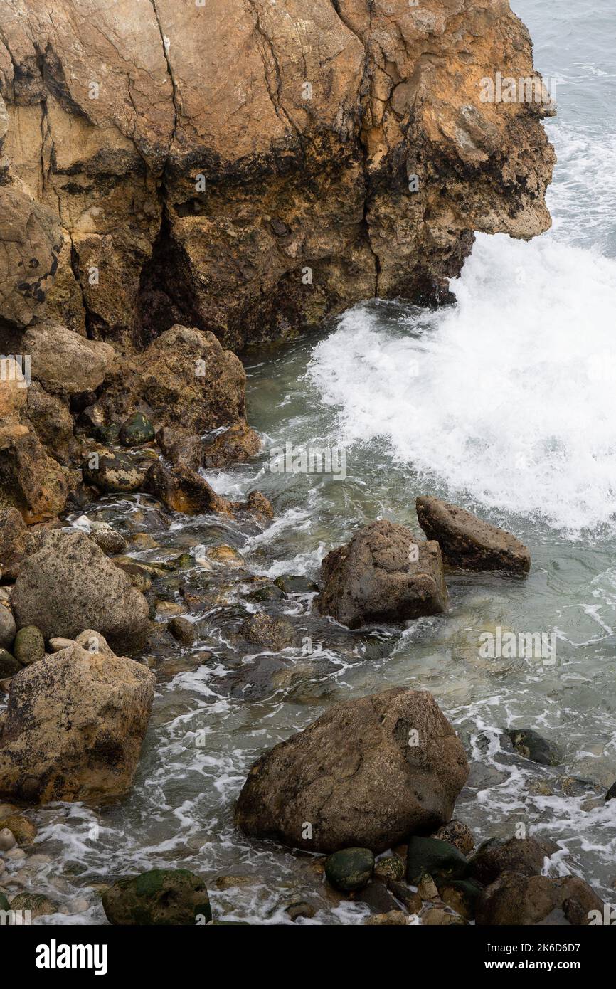 A vertical shot of foamy ocean waves hitting the big coastal rocks ...