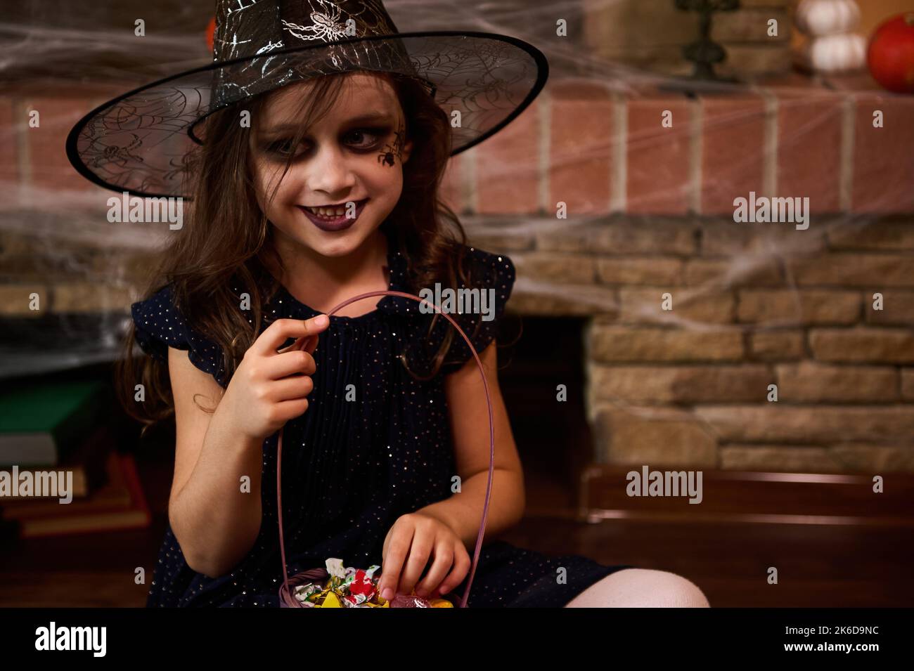 Little girl in witch carnival costume with treat, sitting against a ...