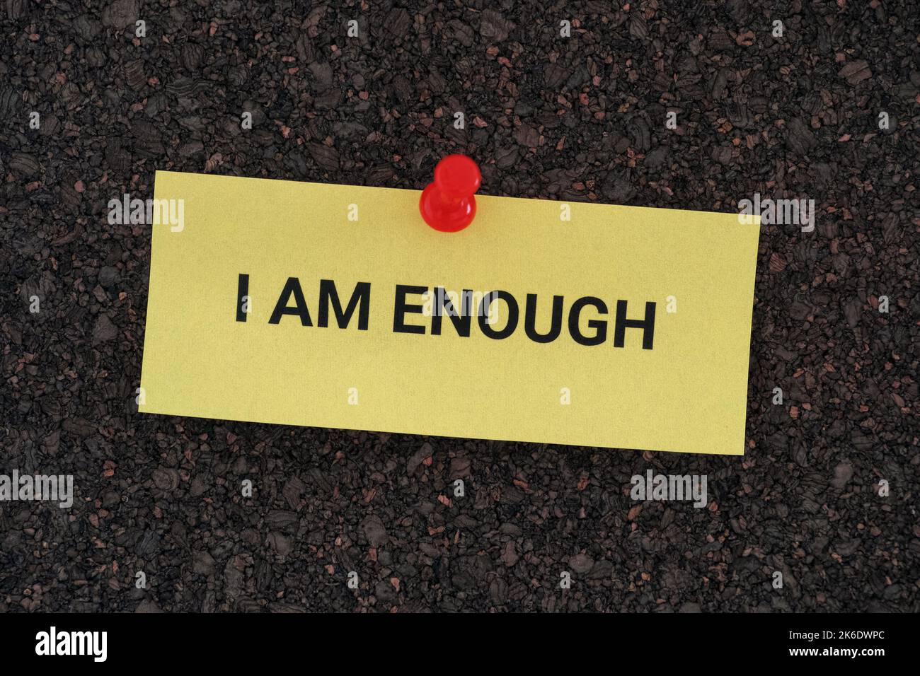 A yellow paper note with the phrase I AM ENOUGH on it pinned to a cork ...