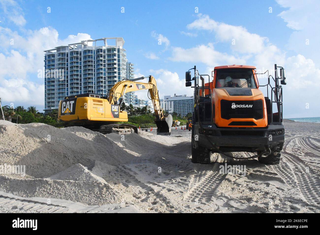 An excavator sits on top a large pile of beach sand and loads sand onto ...