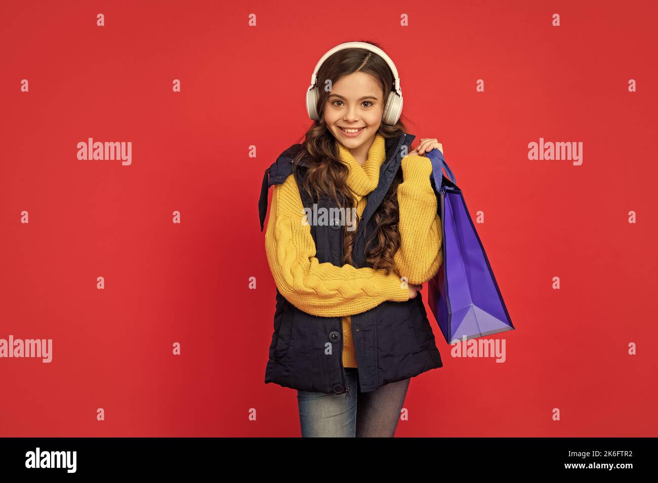 happy kid in winter hat and earphones hold shopping bag on red ...