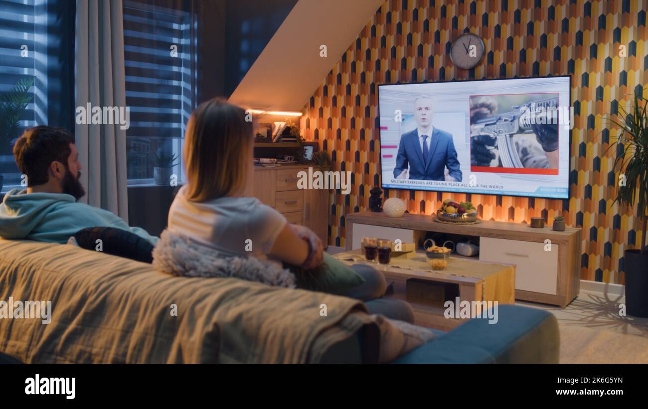 Couple sitting on couch in living room with snacks, watching TV news ...
