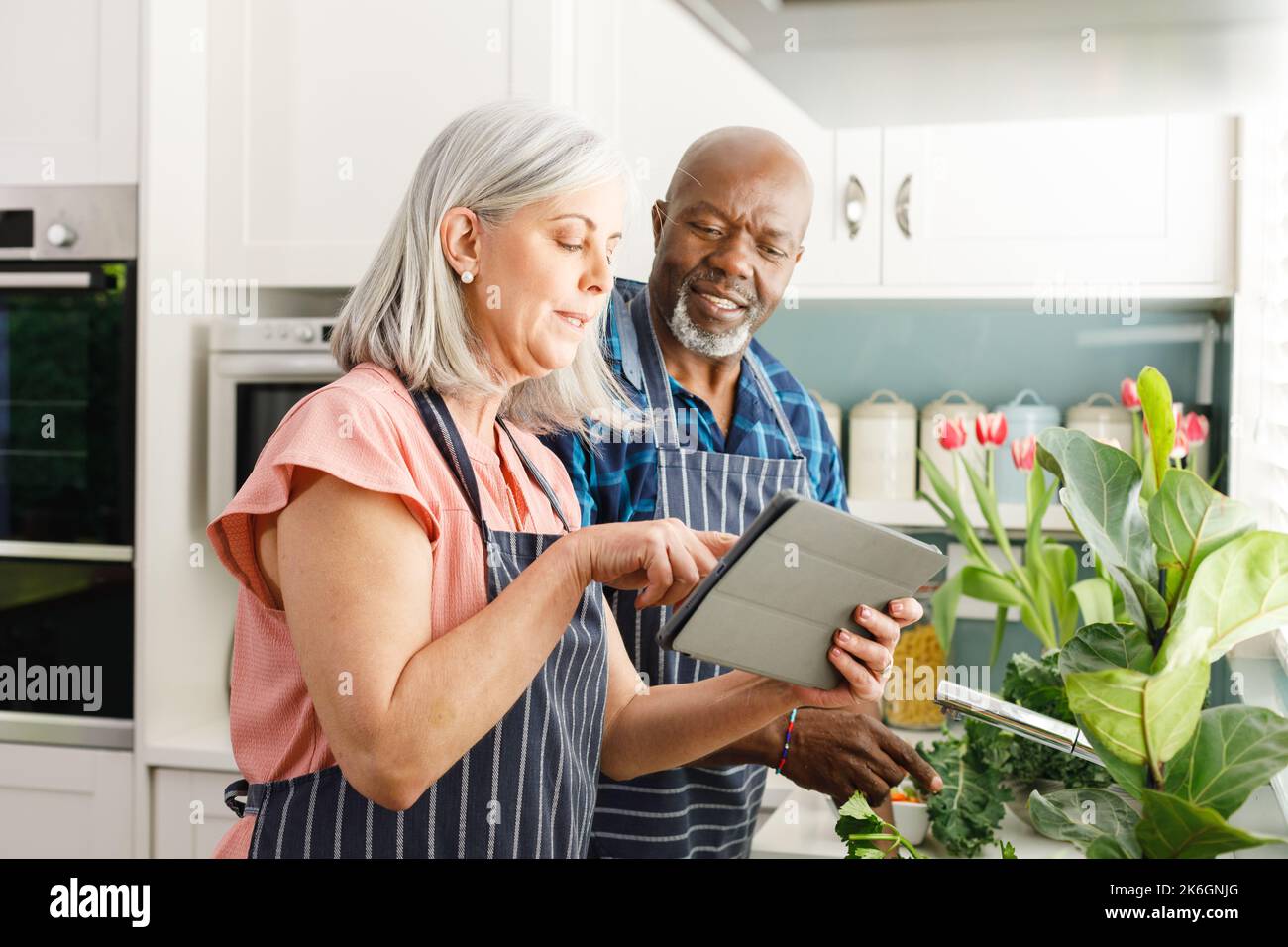 Happy senior diverse couple wearing aprons and using tablet in kitchen ...