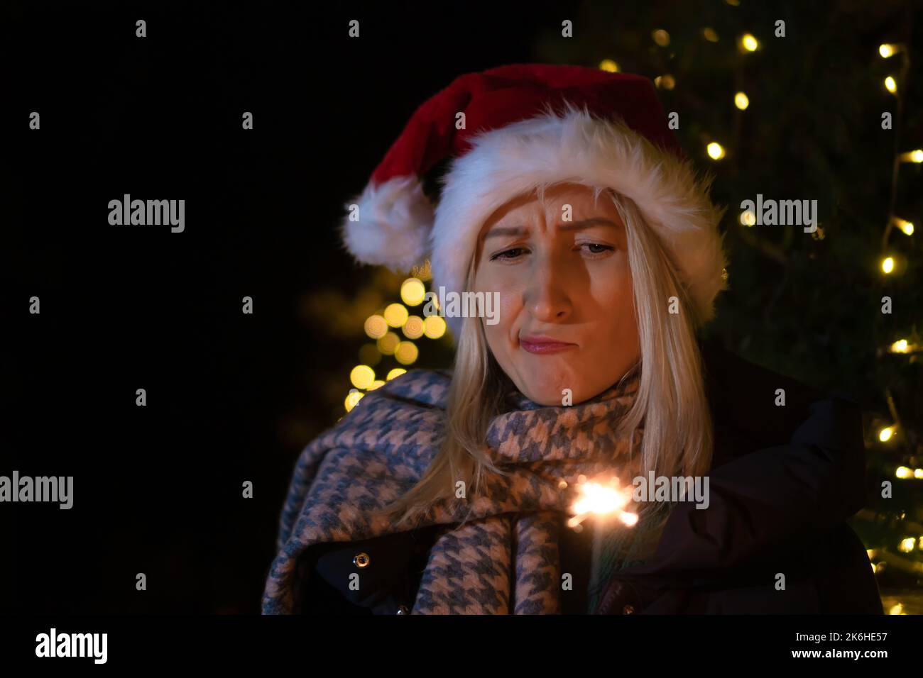 Sad young woman in a Santa Claus hat looks at a sparkler in her hands ...