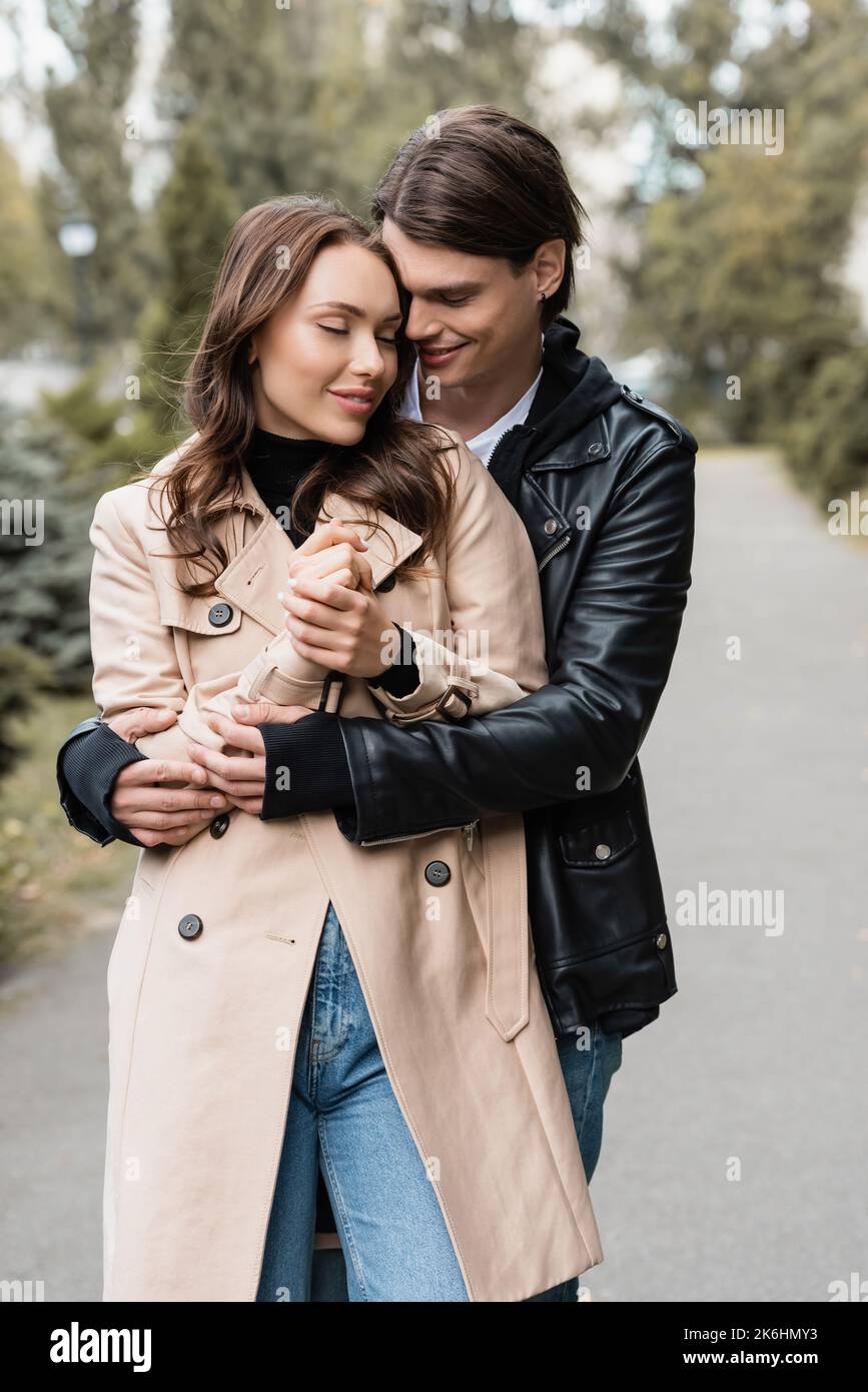 happy young man hugging pleased girlfriend in trench coat outdoors ...