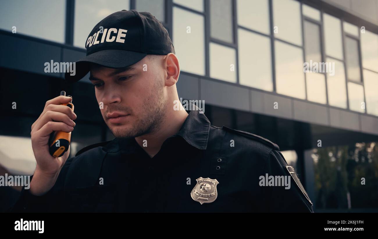 young police officer in uniform and cap using walkie talkie outdoors ...