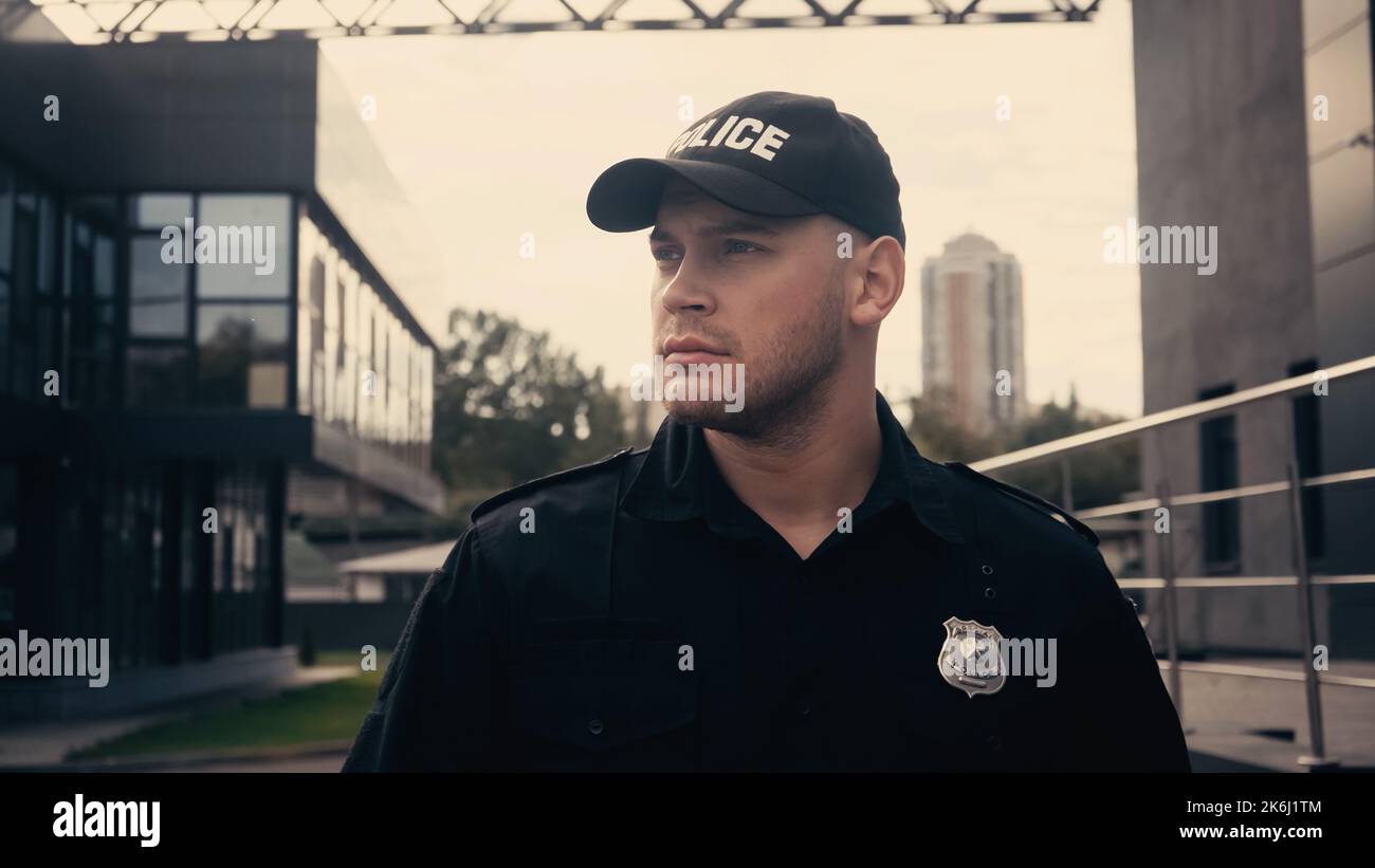 young policeman standing in cap and uniform looking away while ...