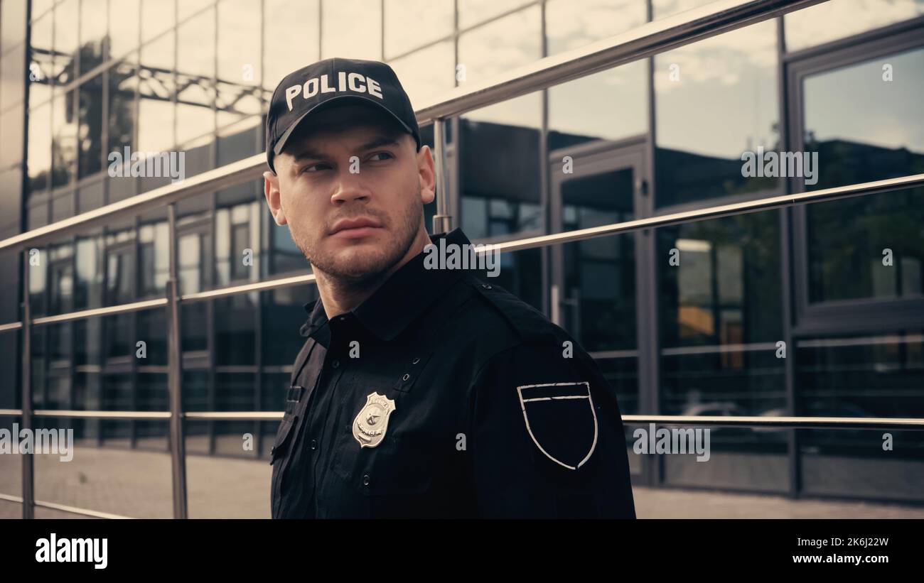 serious policeman standing in cap and uniform while looking away near ...