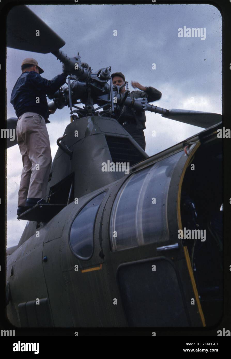 Two technicians, standing atop the helicopter, examine the engine of ...