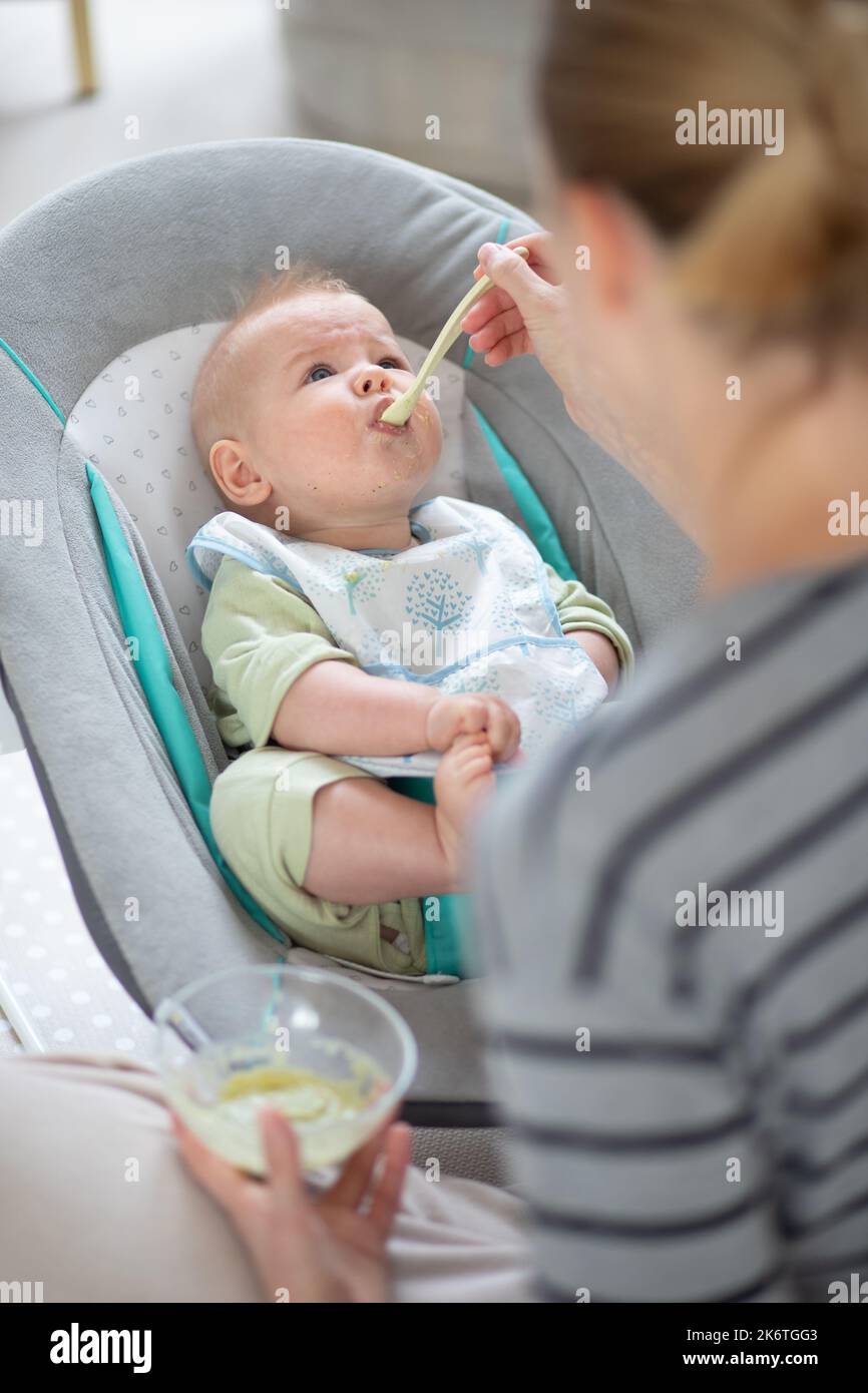 Mother spoon feeding her baby boy infant child in baby chair with fruit ...