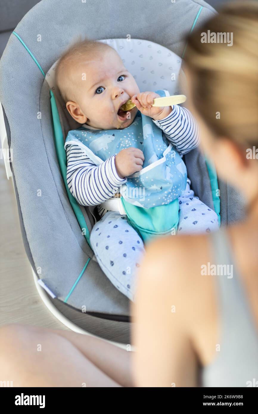 Mother spoon feeding her baby boy infant child in baby chair with fruit ...