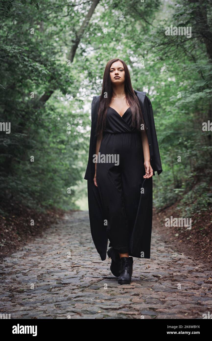 A tall beautiful woman in a black raincoat walks through a dense forest ...