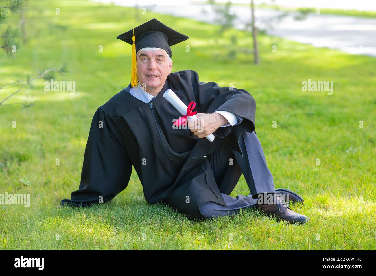 An elderly gray-haired man in a graduation gown sits on green grass ...