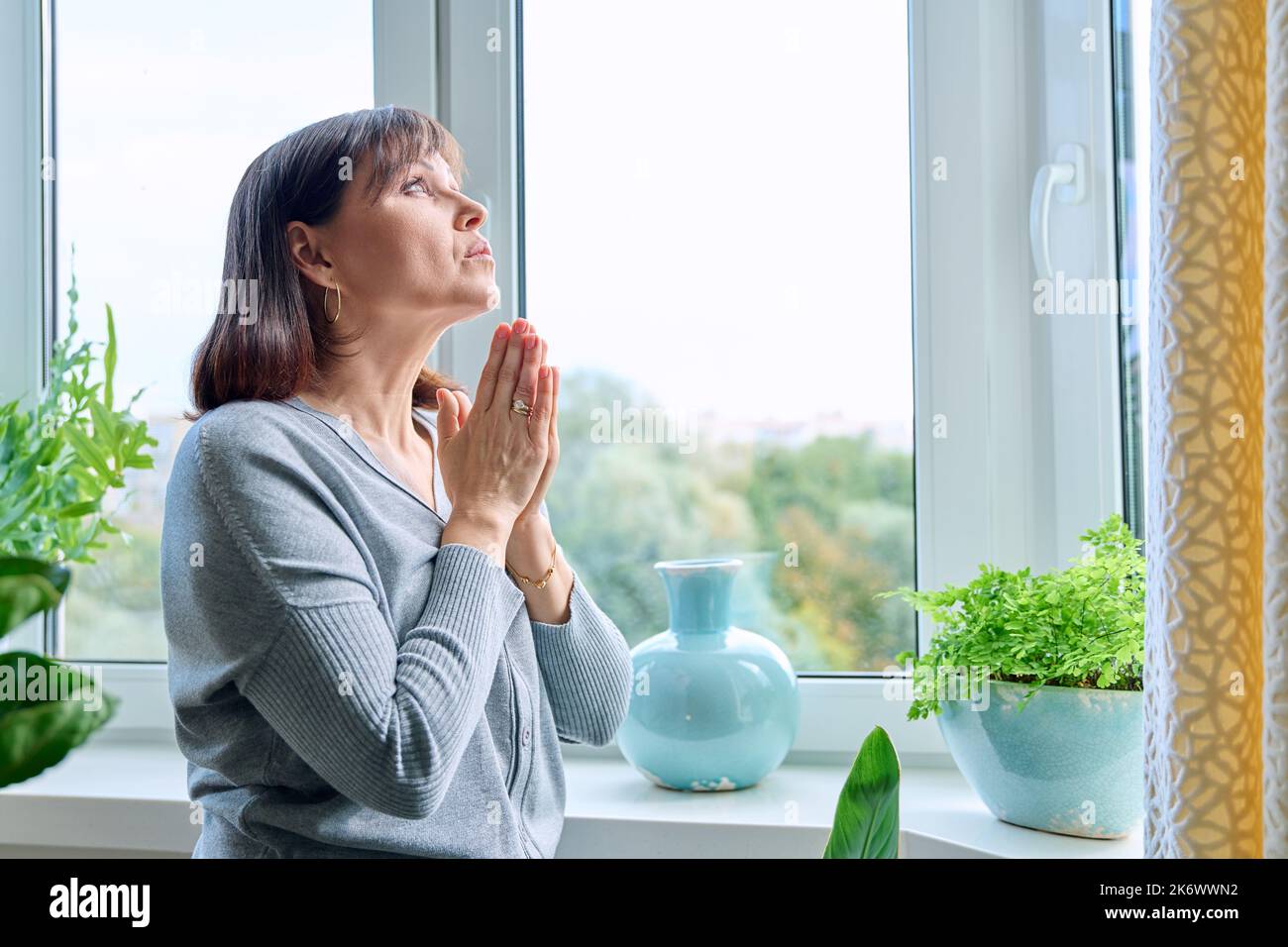 Middle-aged serious woman praying near window at home Stock Photo - Alamy