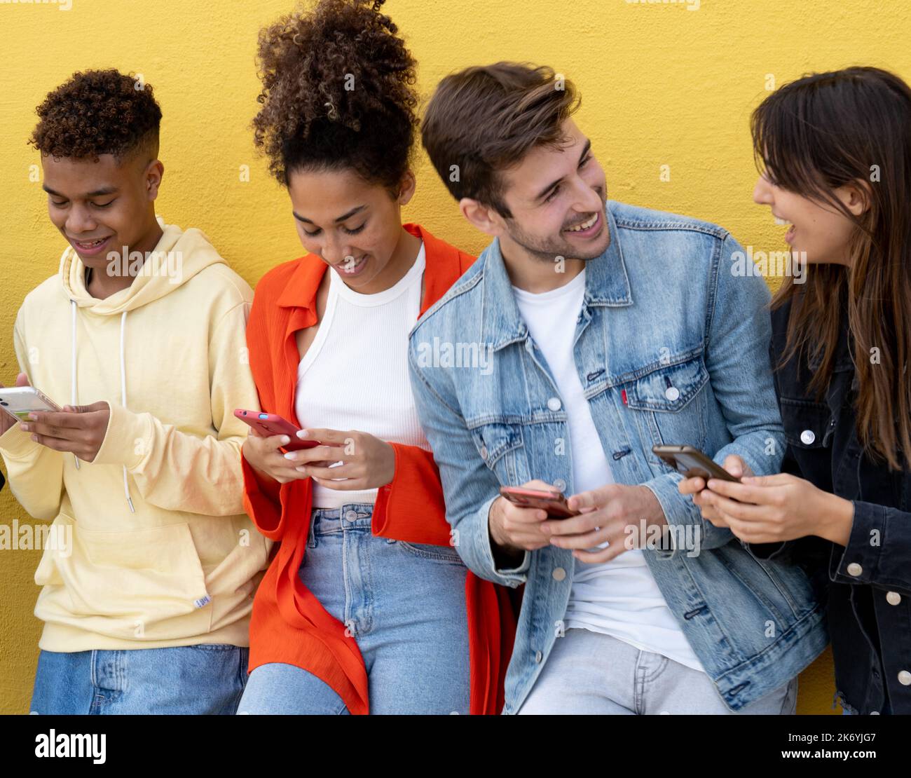 Group of diverse millennial young friends smiling while looking at mobile phone over yellow wall ...