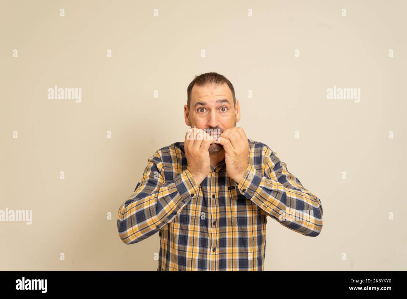 Studio portrait of scared and shocked man in his 40s biting his nails, staring wide-eyed at ...