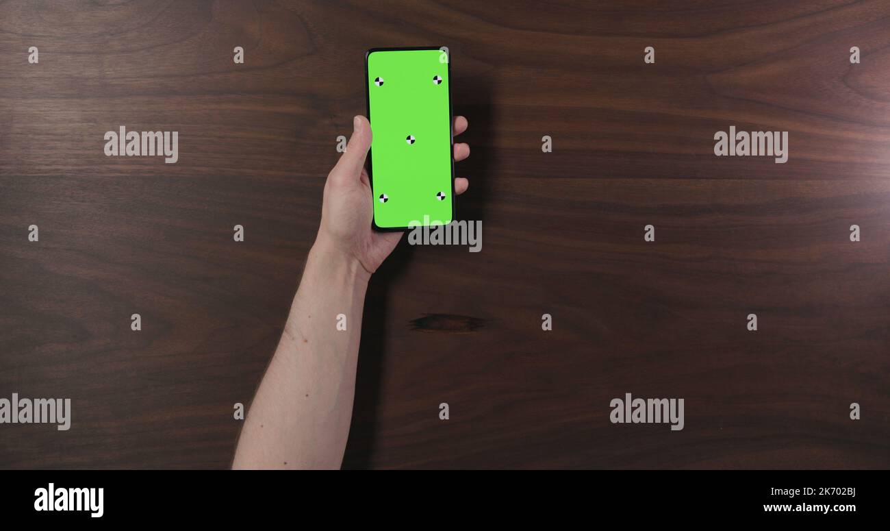 Top view man hand hold smartphone with green screen over black walnut ...