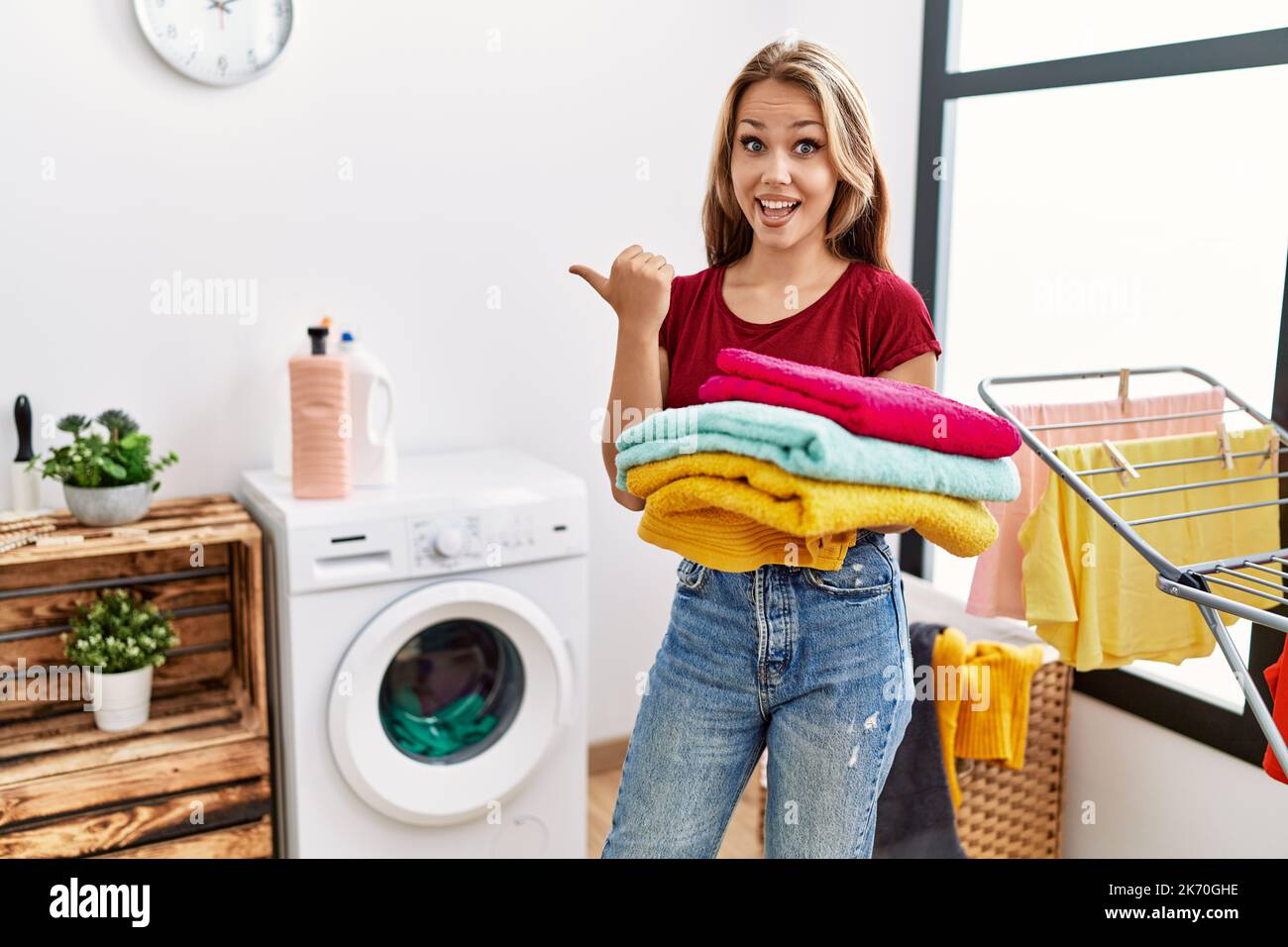 Young caucasian woman holding clean laundry pointing thumb up to the side smiling happy with ...