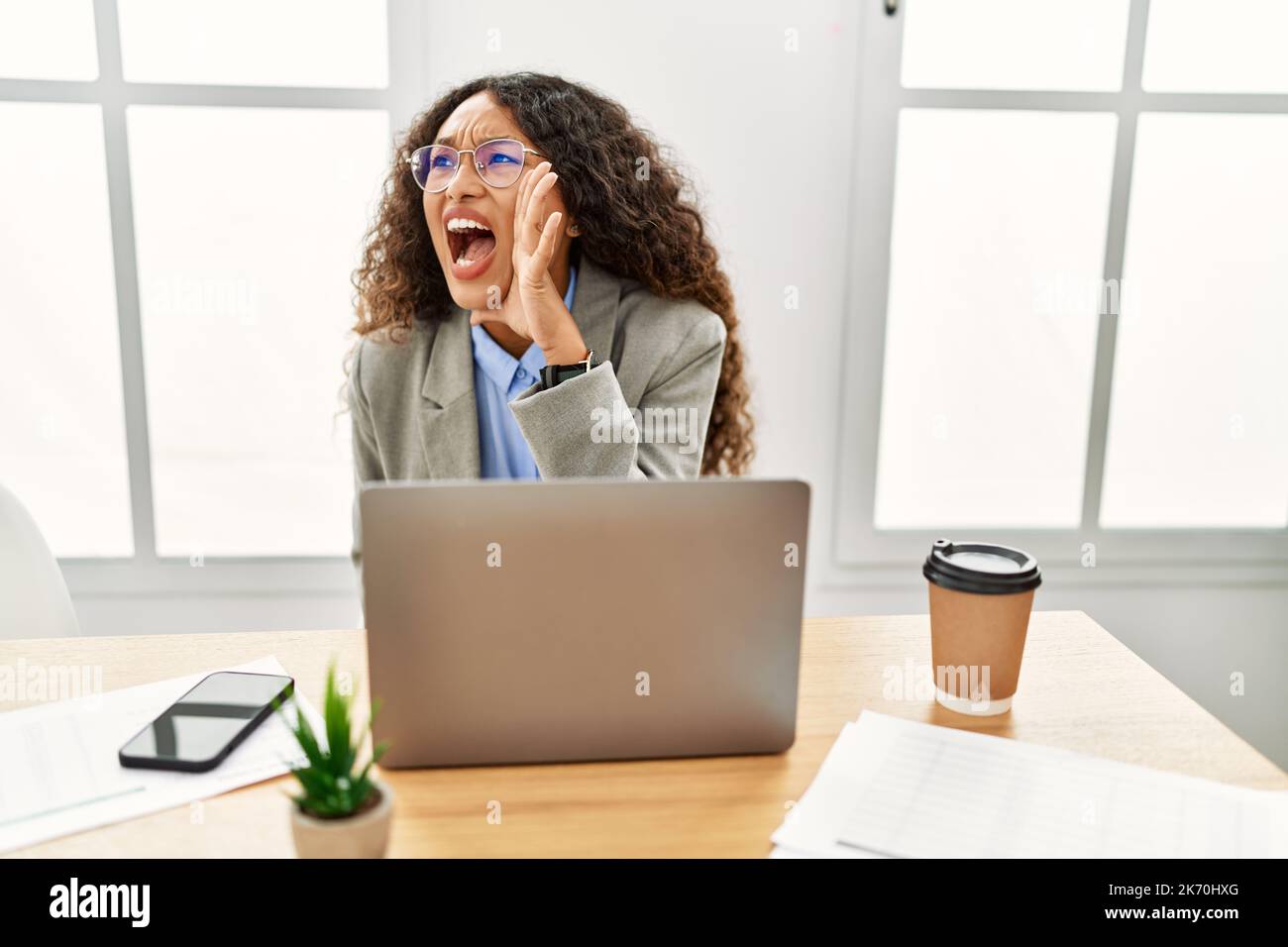 Beautiful hispanic business woman sitting on desk at office working ...