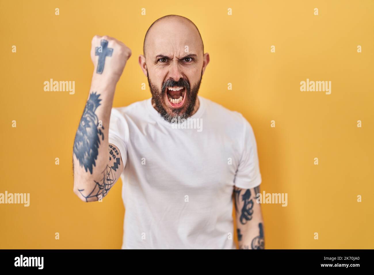 Young hispanic man with tattoos standing over yellow background angry ...