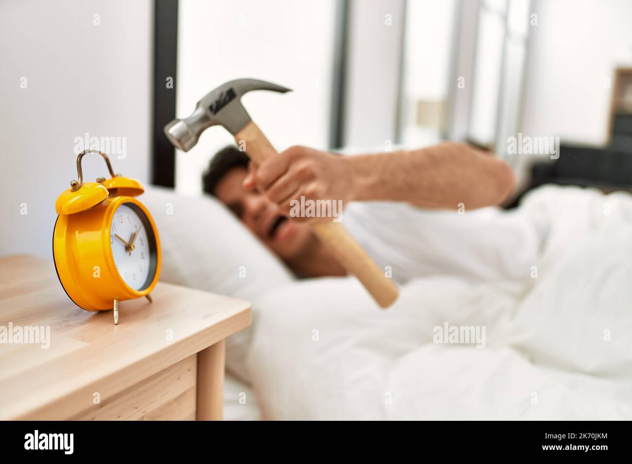 Young Hispanic Man Turning Off Alarm Clock Using Hammer Lying On The