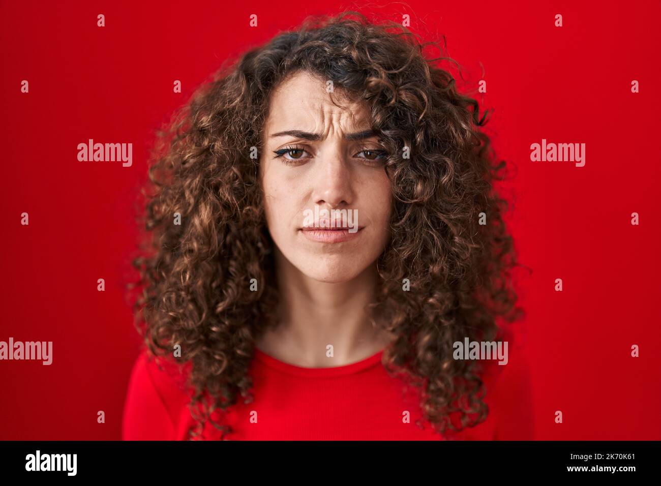 Hispanic woman with curly hair standing over red background depressed ...