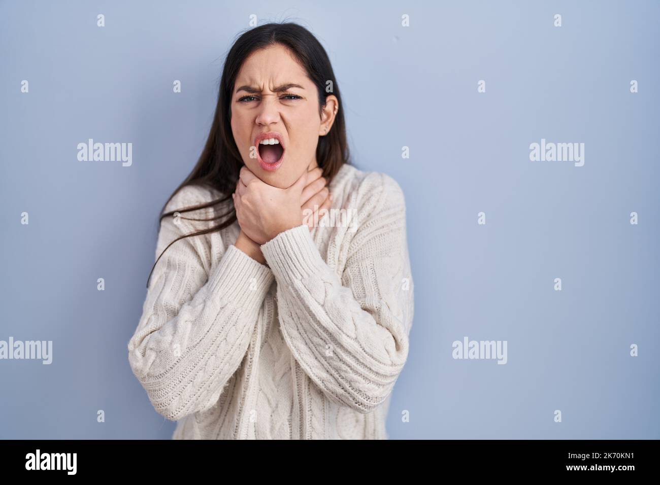 Young brunette woman standing over blue background shouting suffocate ...