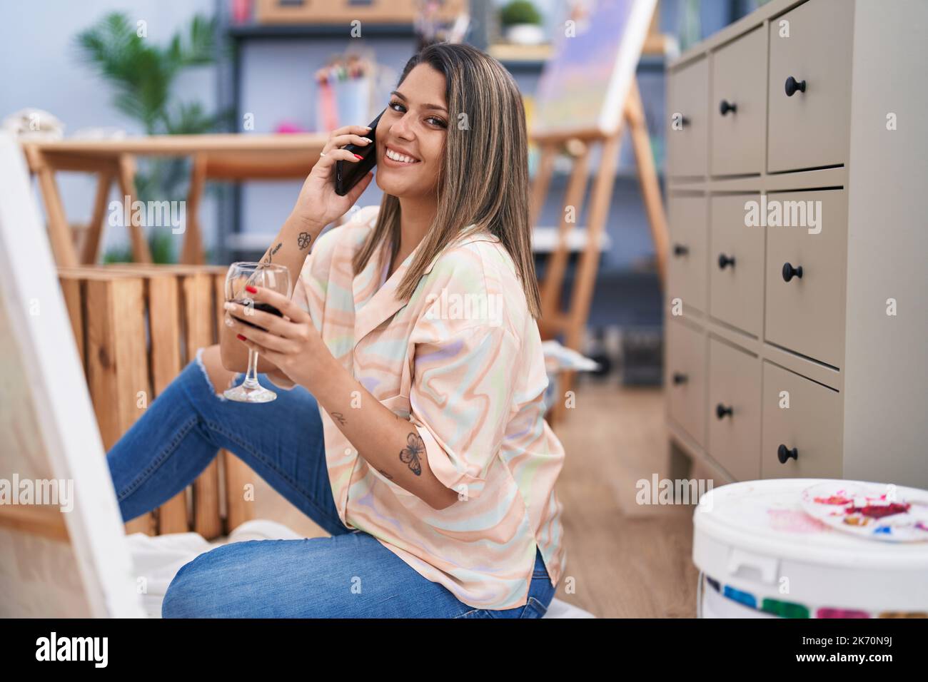 Young hispanic woman artist talking on smartphone drinking wine at art ...