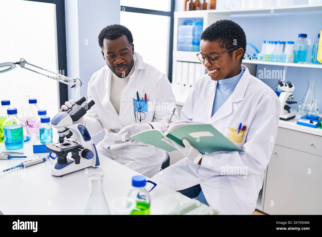 Man and woman scientists using microscope writing on notebook at ...