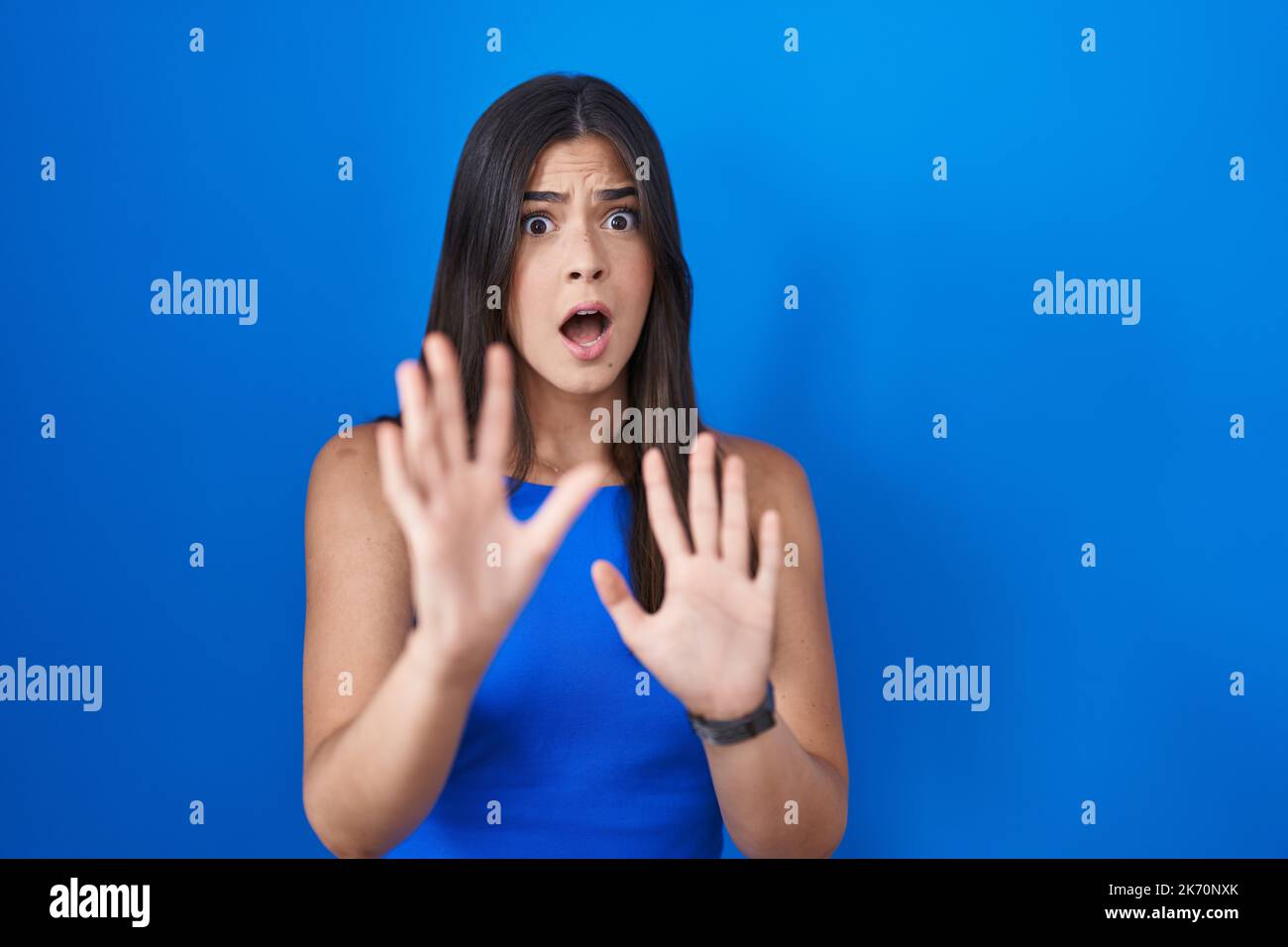 Hispanic woman standing over blue background afraid and terrified with ...